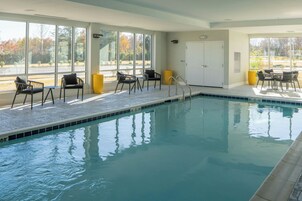 Piscina interior com paredes de vidro transparente, área de estar à beira da piscina com cadeiras e uma porta que dá para outro cômodo.