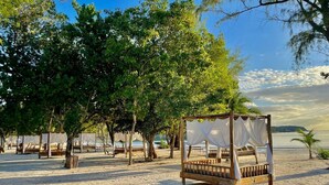 Cenário de praia com cama de madeira sob um toldo, cercada por árvores e céu claro.
