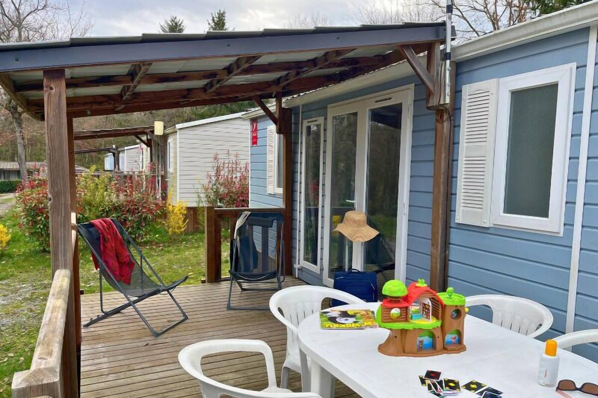 Um deck coberto com mesa e cadeiras, uma pequena área de recreaç ão infantil com brinquedos, e um alpendre coberto com uma cadeira e um casaco vermelho pendurado no corrimão.