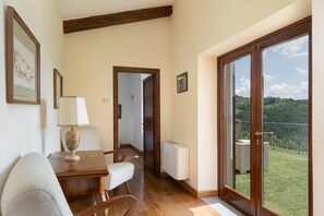 Quarto com chão de madeira, uma cadeira branca, uma mesa com abajur, radiador e porta que dá para uma varanda com vista para o verde.