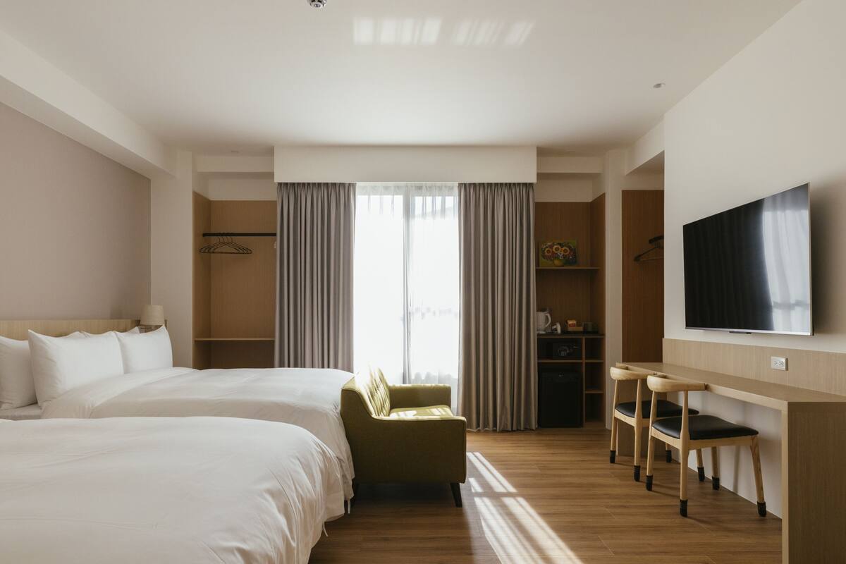 Quarto de hotel com duas camas, uma escrivaninha, televisão e janela com cortinas.