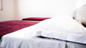 Uma cama com roupa de cama branca e vermelha, uma parede simples e uma janela ao fundo.