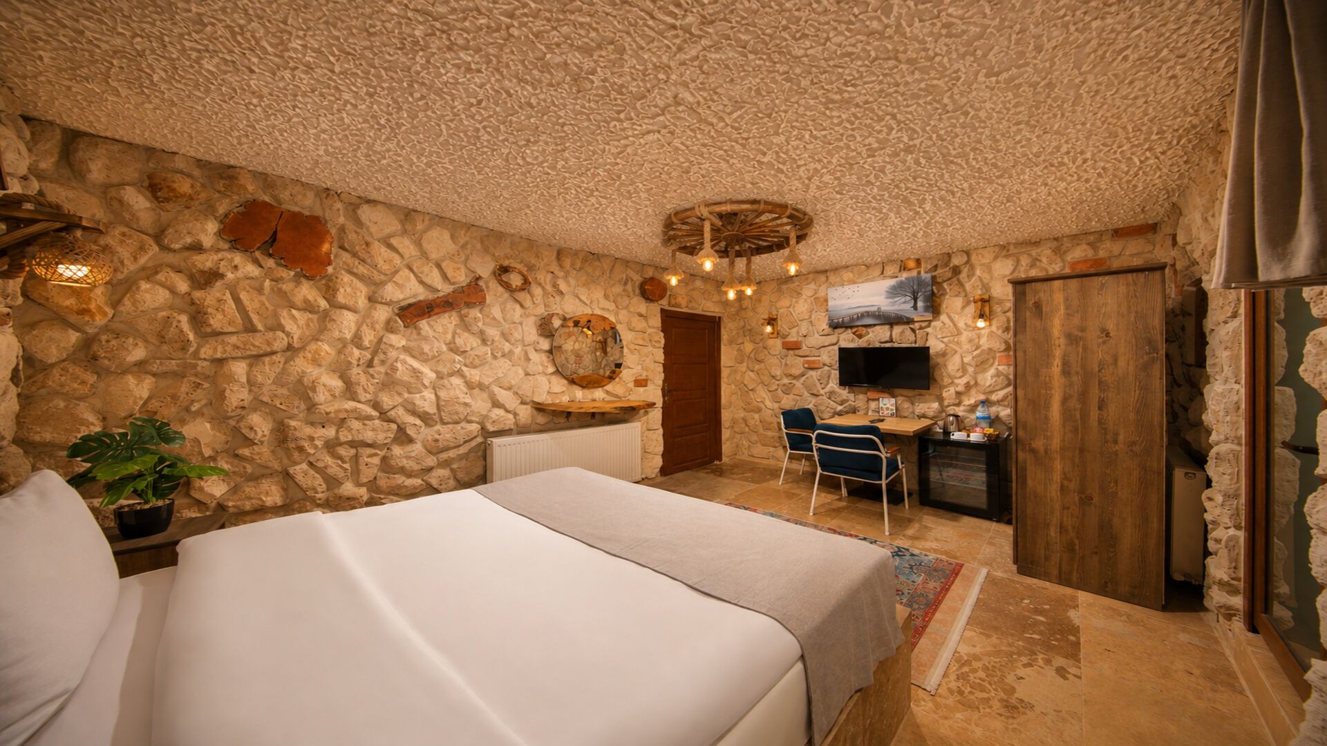 Quarto com paredes de pedra, uma cama, uma escrivaninha e uma televisão.