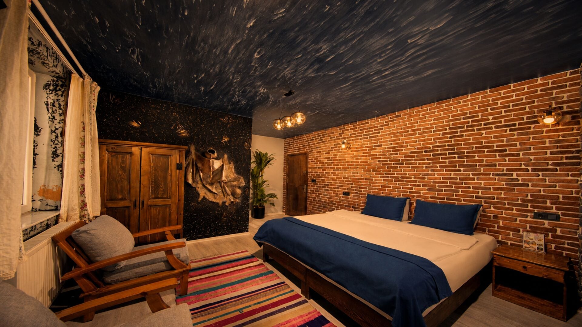 Quarto com parede de tijolos, cama com roupa de cama azul, guarda-roupa de madeira e um tapete listrado e colorido.