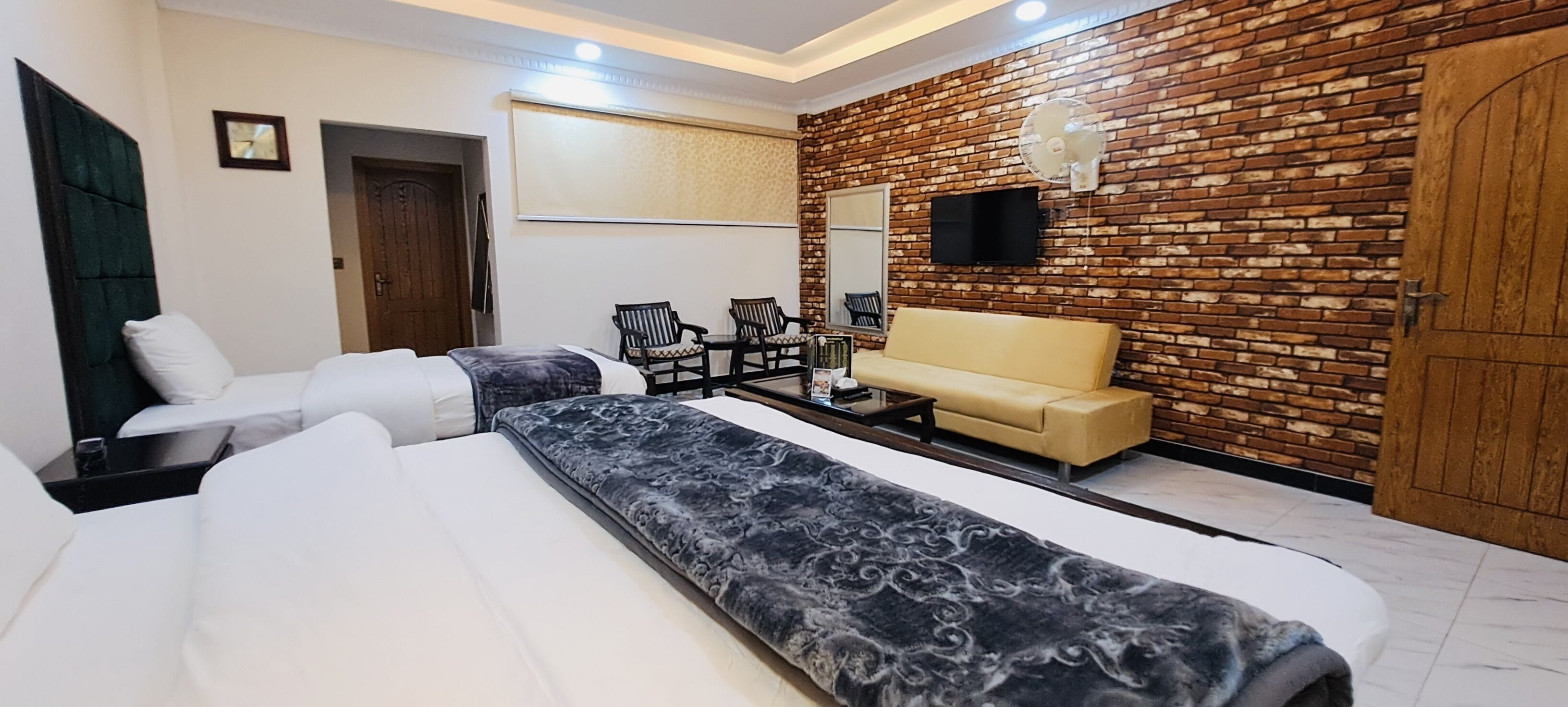 Quarto de hotel moderno com parede em padrão tijolo, um sofá, duas camas de solteiro, uma TV e um ventilador.