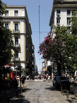 Rua com edifícios, uma rotatória e pedestres.