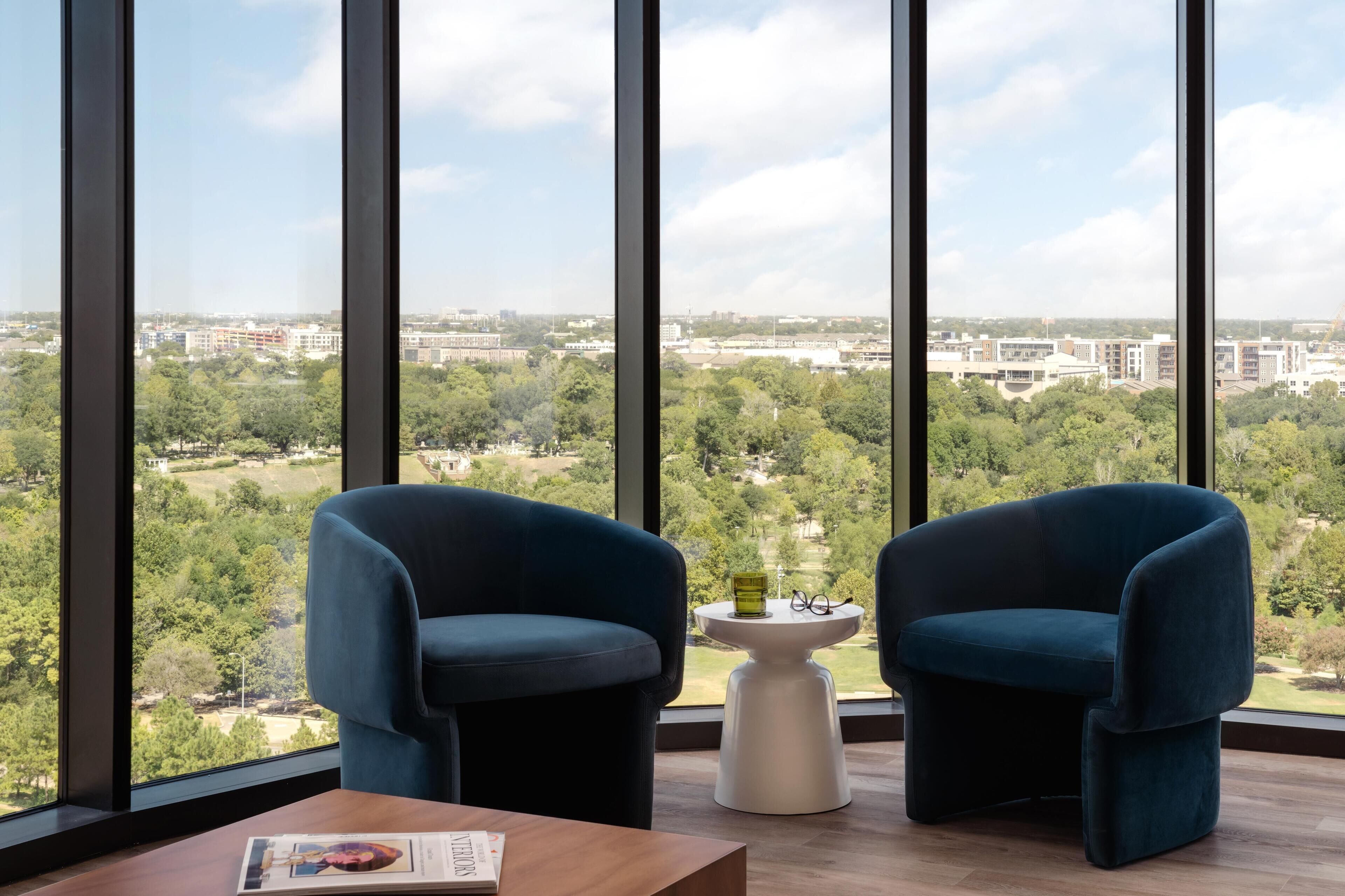 Um quarto com duas poltronas azul-escuro voltadas para uma janela ampla com vista para a cidade e áreas verdes.