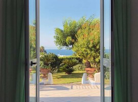 Visão através de uma porta de vidro de um jardim com um espaço de pátio, um vaso grande e vista para o mar.