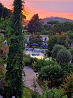 Um jardim com piscina, rodeado por vegetação exuberante e um pôr do sol ao fundo.