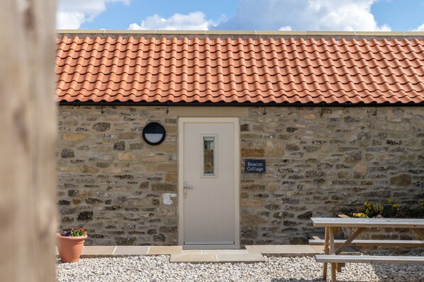Um edifício de pedra com telhado de telhas, uma porta e uma placa com a inscrição 'Beacon Cottage'.