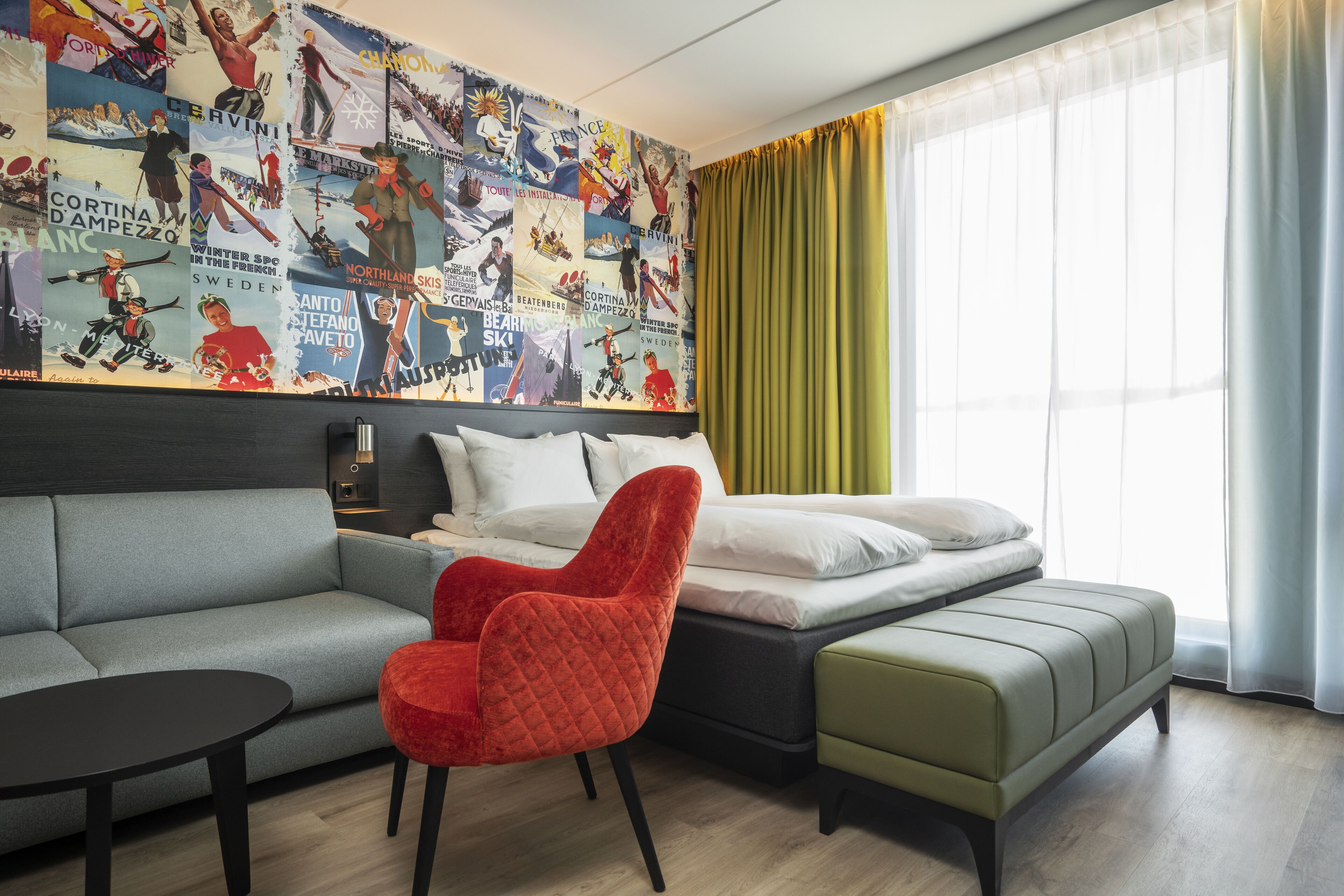 Quarto de hotel com uma cama, uma cadeira vermelha, um sofá cinzento, uma mesinha e um painel decorado com vários cartazes de desporto.