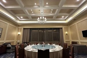 Uma sala de conferências com uma mesa redonda, cadeiras, um lustre e uma televisão na parede.