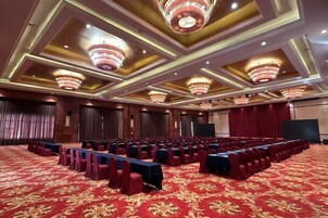 Sala de conferências com fileiras de mesas e cadeiras, grandes lustres e piso coberto por um tapete vermelho.