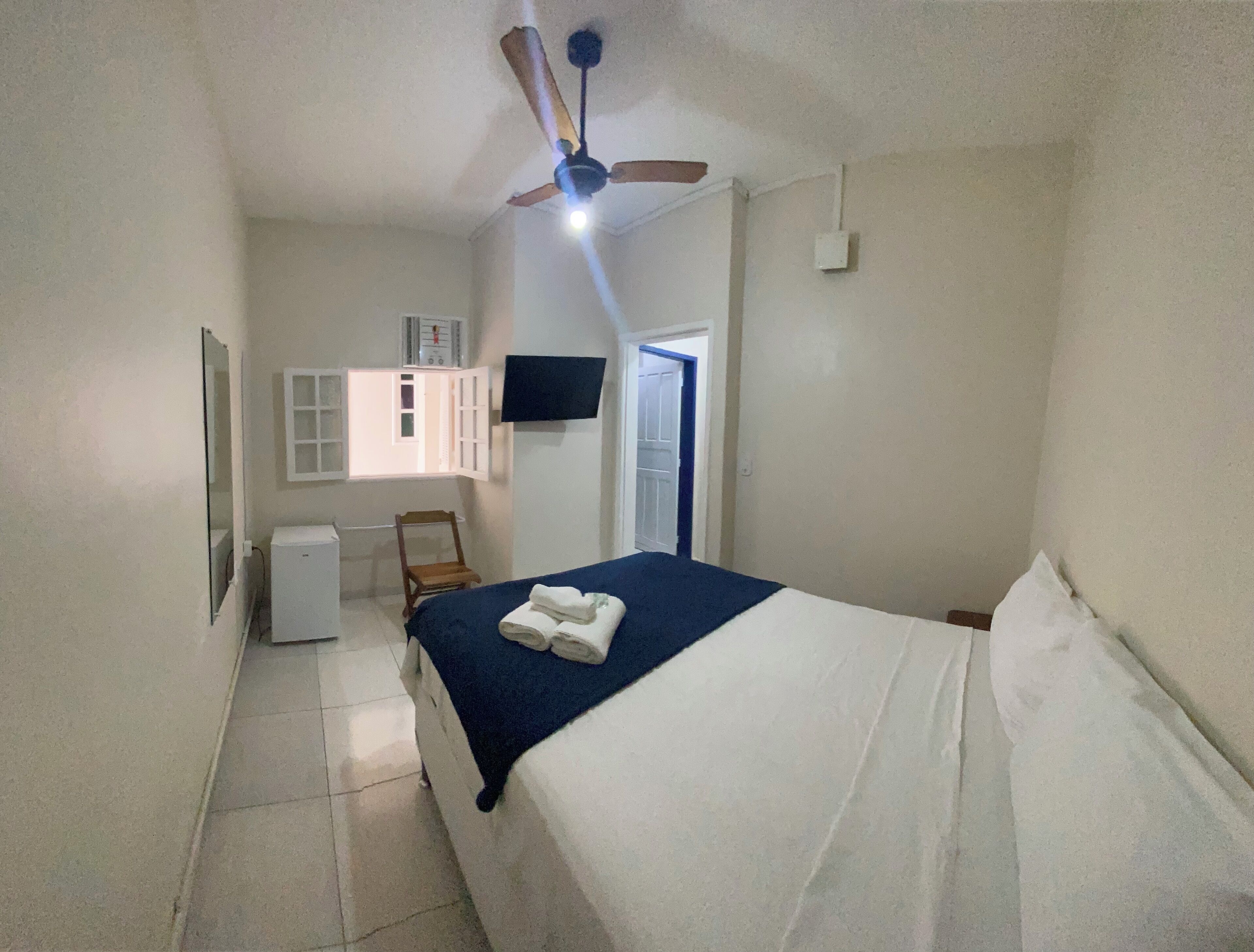 Quarto pequeno e limpo com cama, cadeira, escrivaninha, televisão e ventoinha no teto.