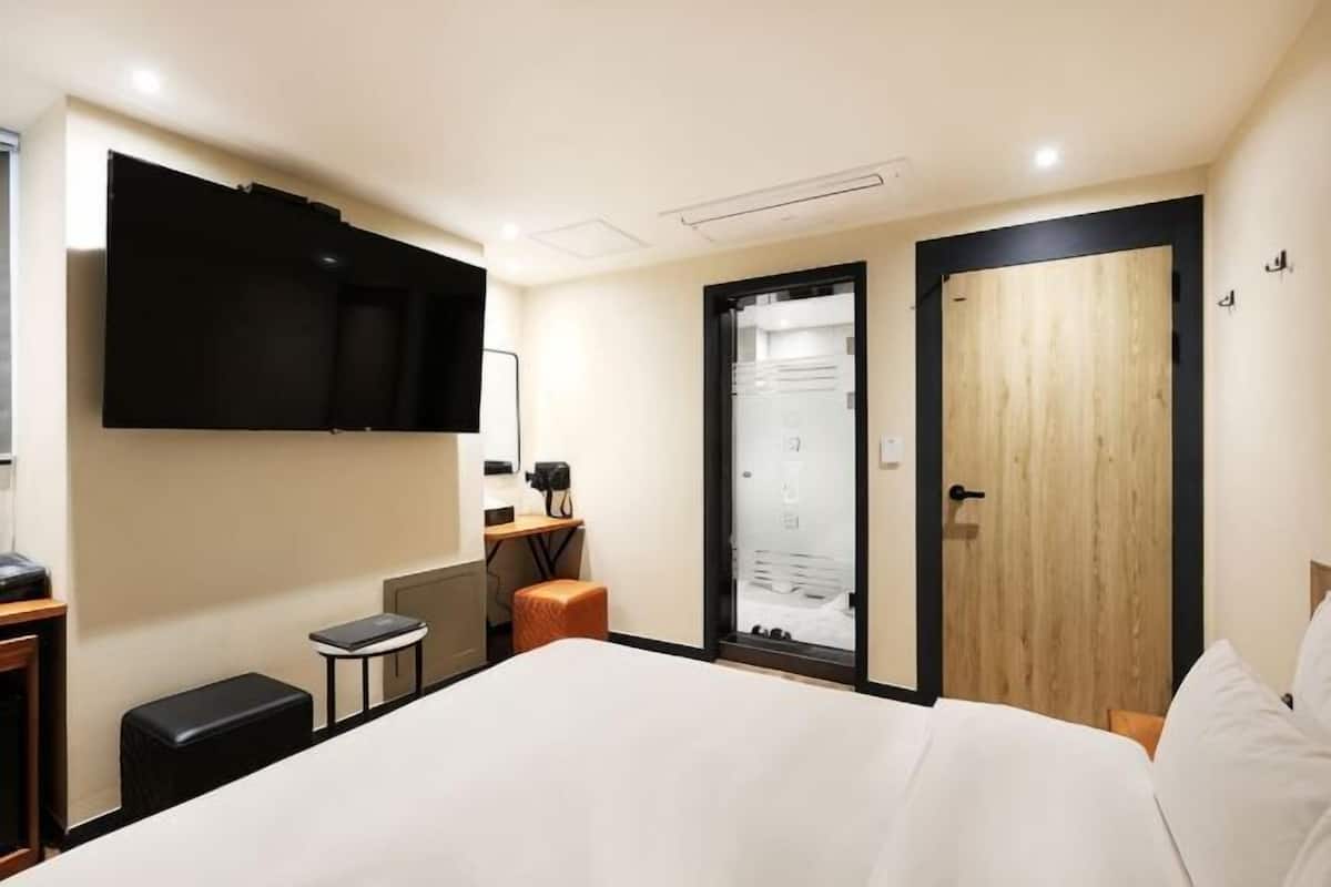 Quarto de hotel com uma cama, uma escrivaninha com computador, uma TV fixada na parede e uma porta de madeira.