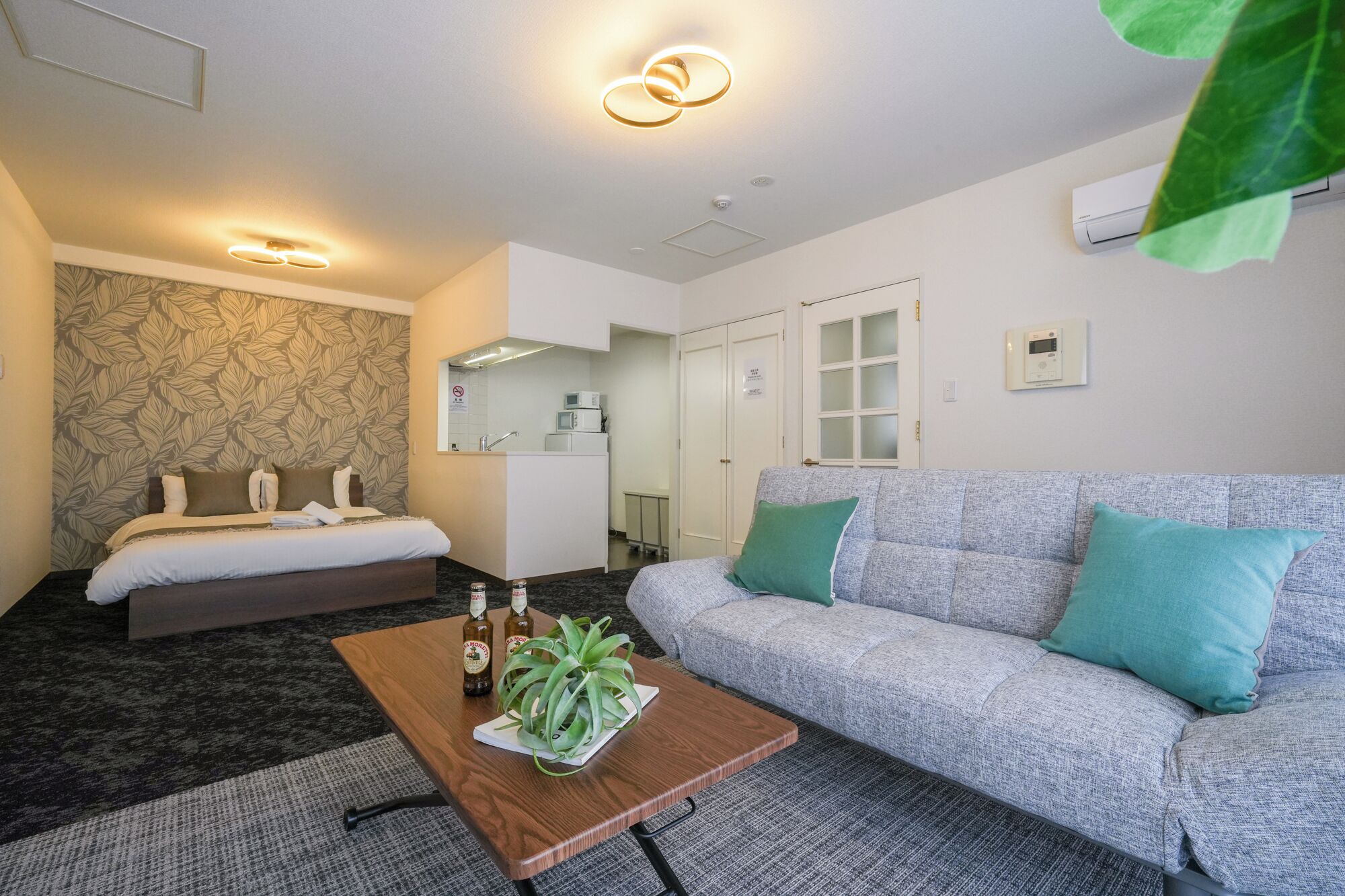 Quarto de hotel com cama, sofá, mesa de centro com garrafas e planta, área de cozinha e banheiro.