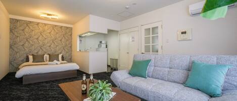 Quarto de hotel com cama, sofá, mesa de centro com garrafas e planta, área de cozinha e banheiro.
