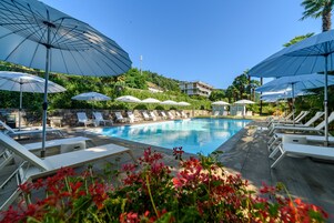 Área de piscina com espreguiçadeiras e guarda-sóis, cercada por vegetação e uma encosta.