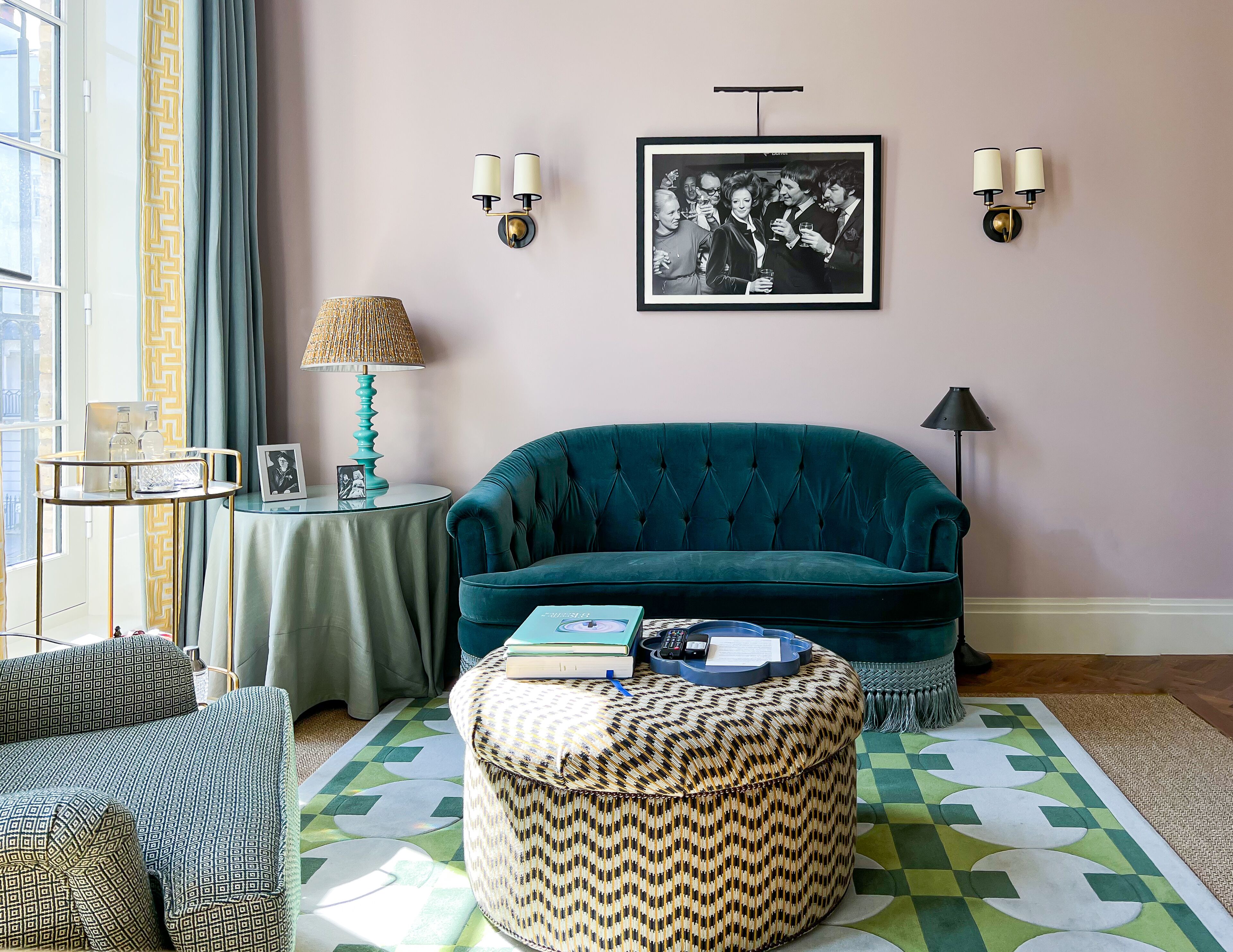 Uma sala de estar com um sofá verde-água, um banquinho estampado, uma pequena mesa lateral com abajur e uma fotografia em preto e branco emoldurada na parede.
