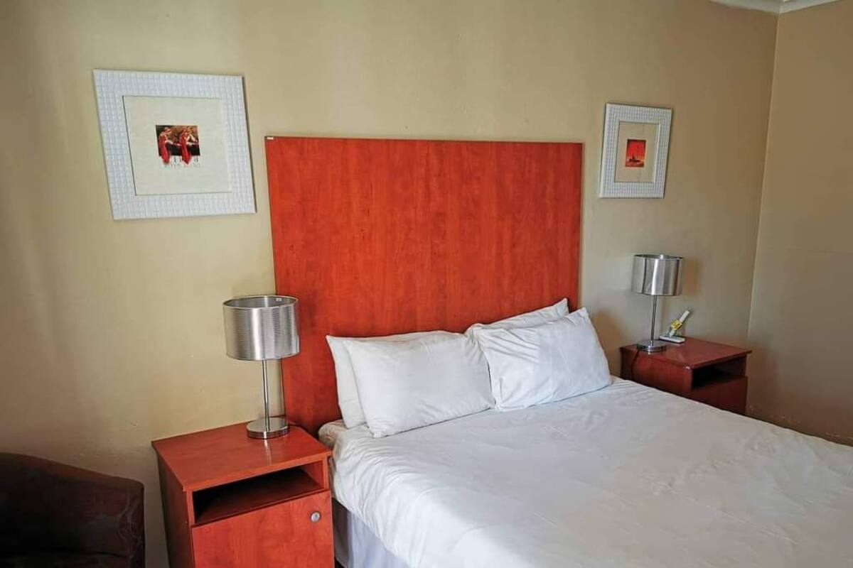 Quarto de hotel com cabeceira de madeira, cama com roupa de cama branca, duas mesas de cabeceira com abajures e quadros nas paredes.