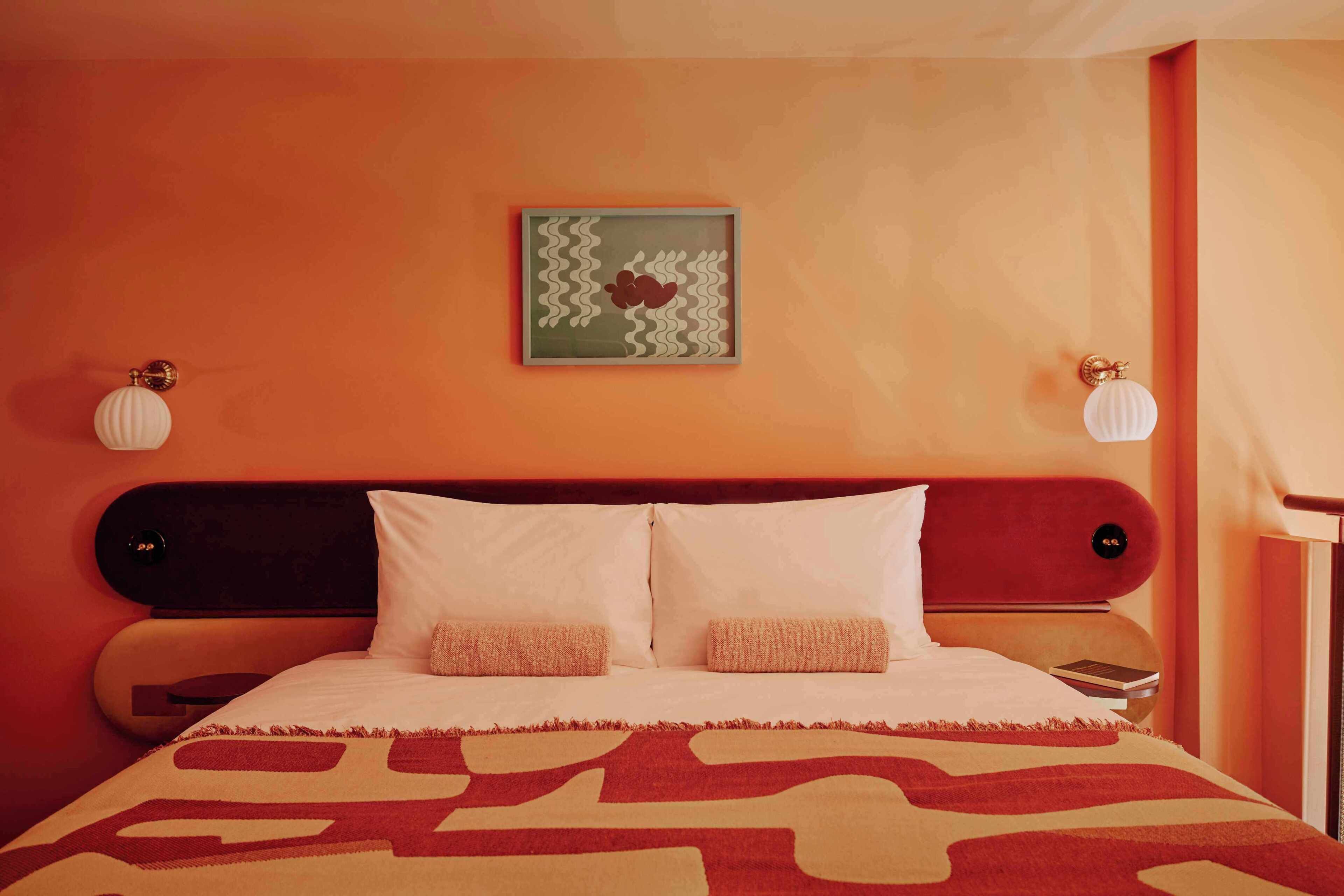 Quarto de hotel com uma cama, uma mesinha de cabeceira, uma luminária na parede e uma obra de arte emoldurada.
