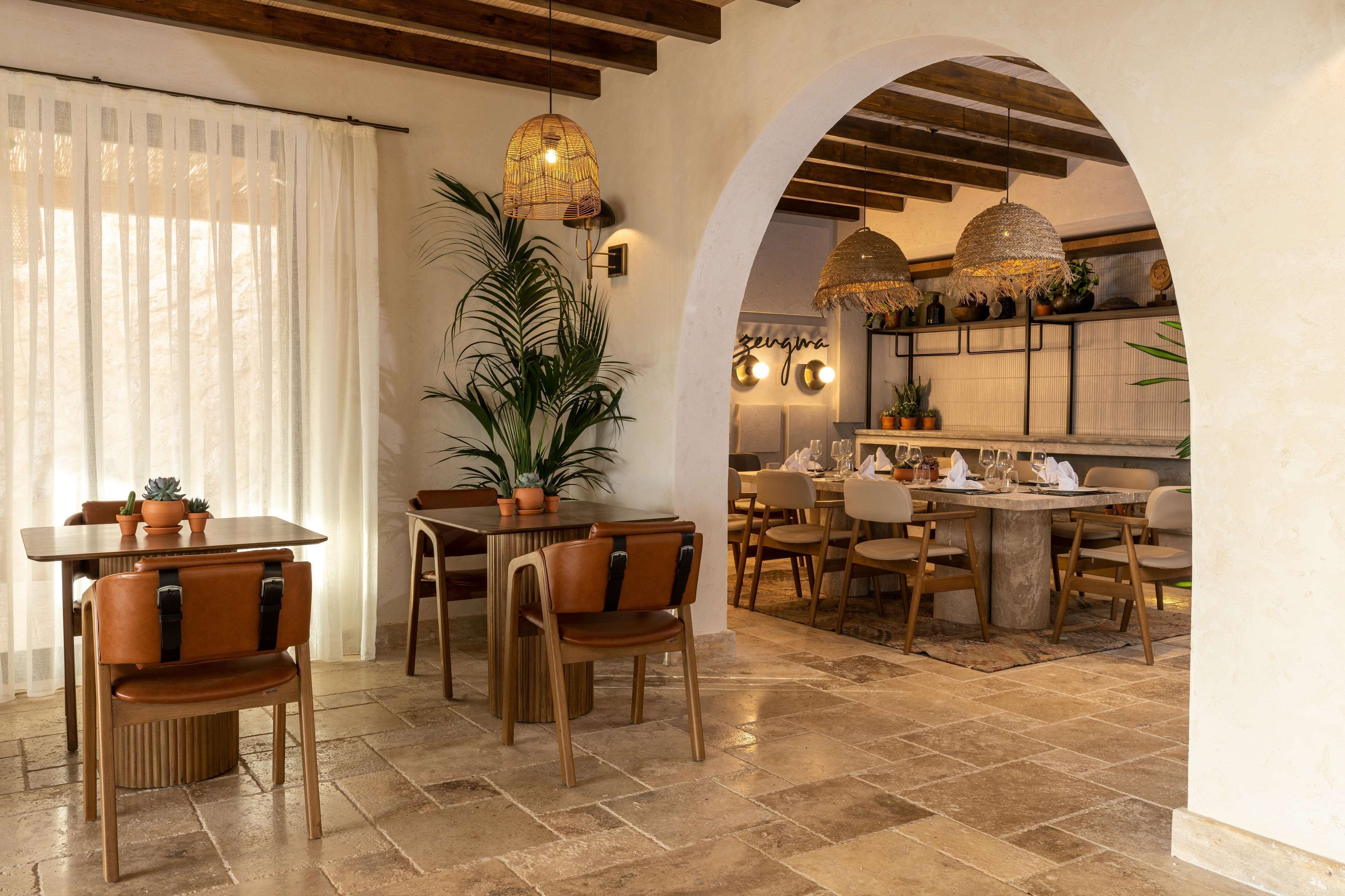 Uma sala de jantar com cadeiras e mesas de madeira, uma planta grande em vaso e luminárias de vime suspensas.