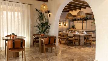 Uma sala de jantar com cadeiras e mesas de madeira, uma planta grande em vaso e luminárias de vime suspensas.