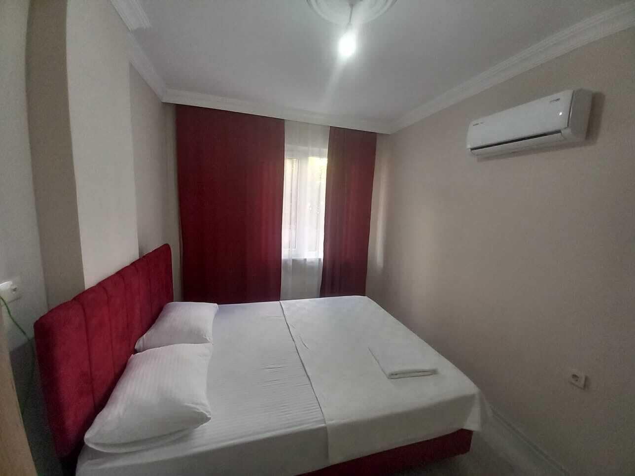 Quarto de hotel com uma cama de solteiro, cortinas vermelhas, luminária de teto branca e um aparelho de ar condicionado na parede.