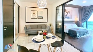 Quarto de hotel moderno com sofá cinzento, mesa de jantar com cadeiras, cama com colcha azul e uma janela grande com cortinas.