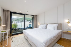 Quarto de hotel moderno com uma cama grande, uma escrivaninha, uma cadeira e vista para a cidade através de amplas janelas.