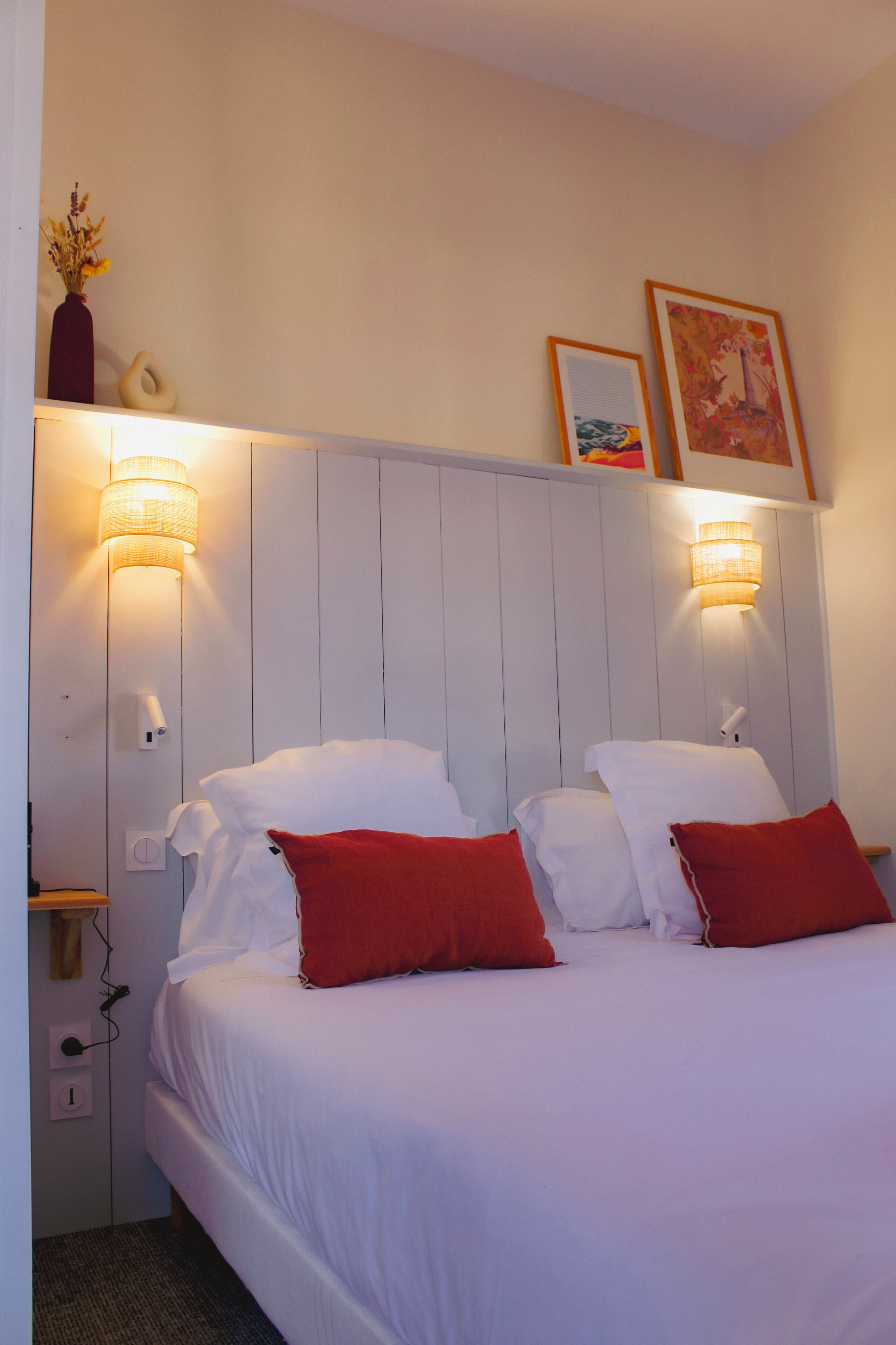 Quarto de hotel com uma cama, duas luminárias fixadas na parede, um vaso com flores secas e duas fotografias emolduradas na parede.