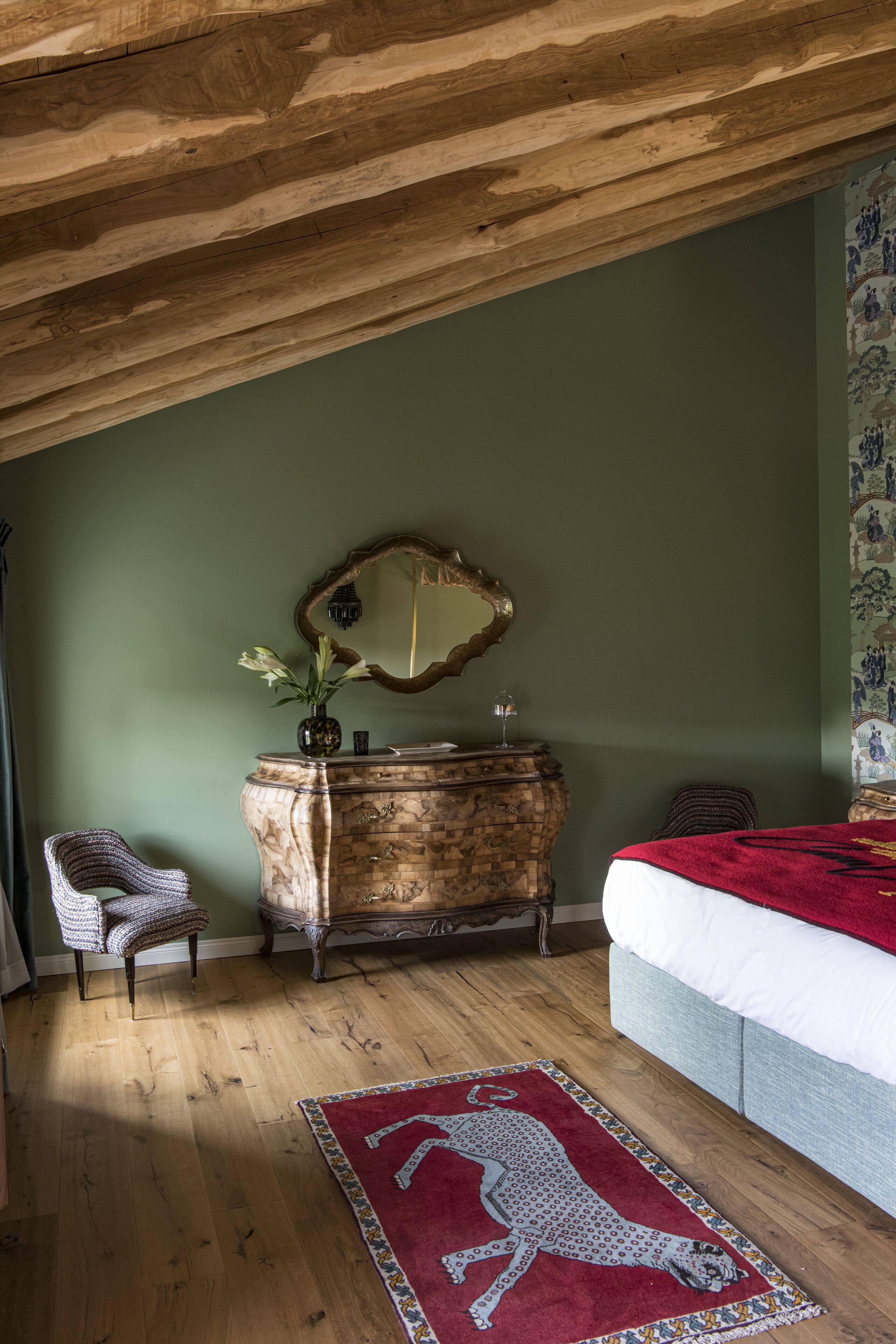 Um quarto com teto de madeira, uma cama com cobertor vermelho, cômoda com espelho e tapete estampado.