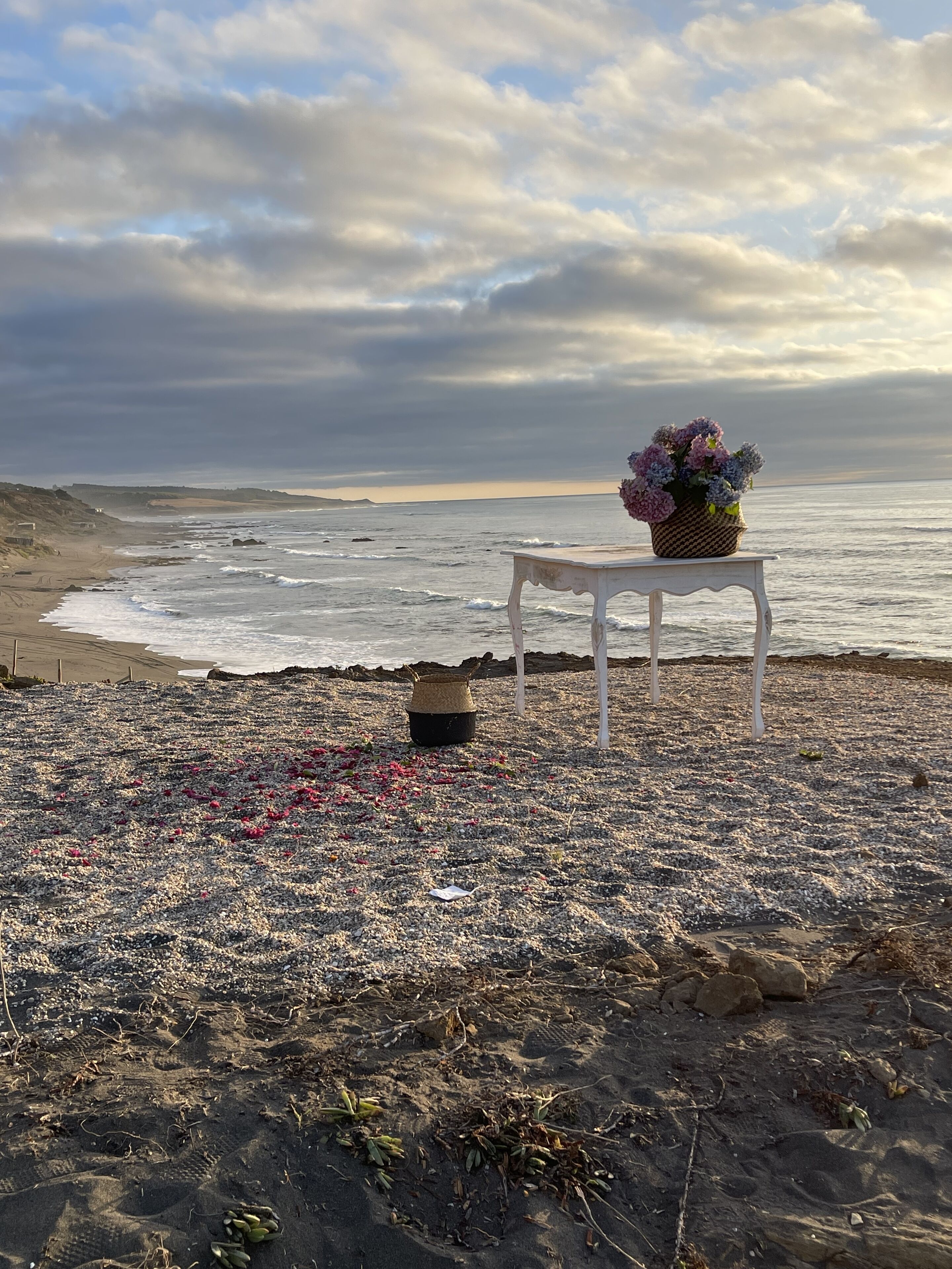 Uma praia com uma mesa branca e um cesto de flores, cercada por destroços e pedras espalhadas.