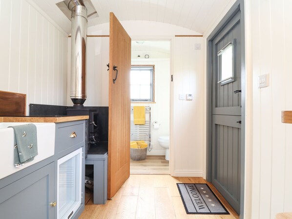 Um corredor estreito com porta de madeira, fogão de metal e um pequeno banheiro visível através de uma porta aberta.
