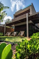 Uma casa moderna com varanda coberta, duas poltronas de descanso e vegetação exuberante.