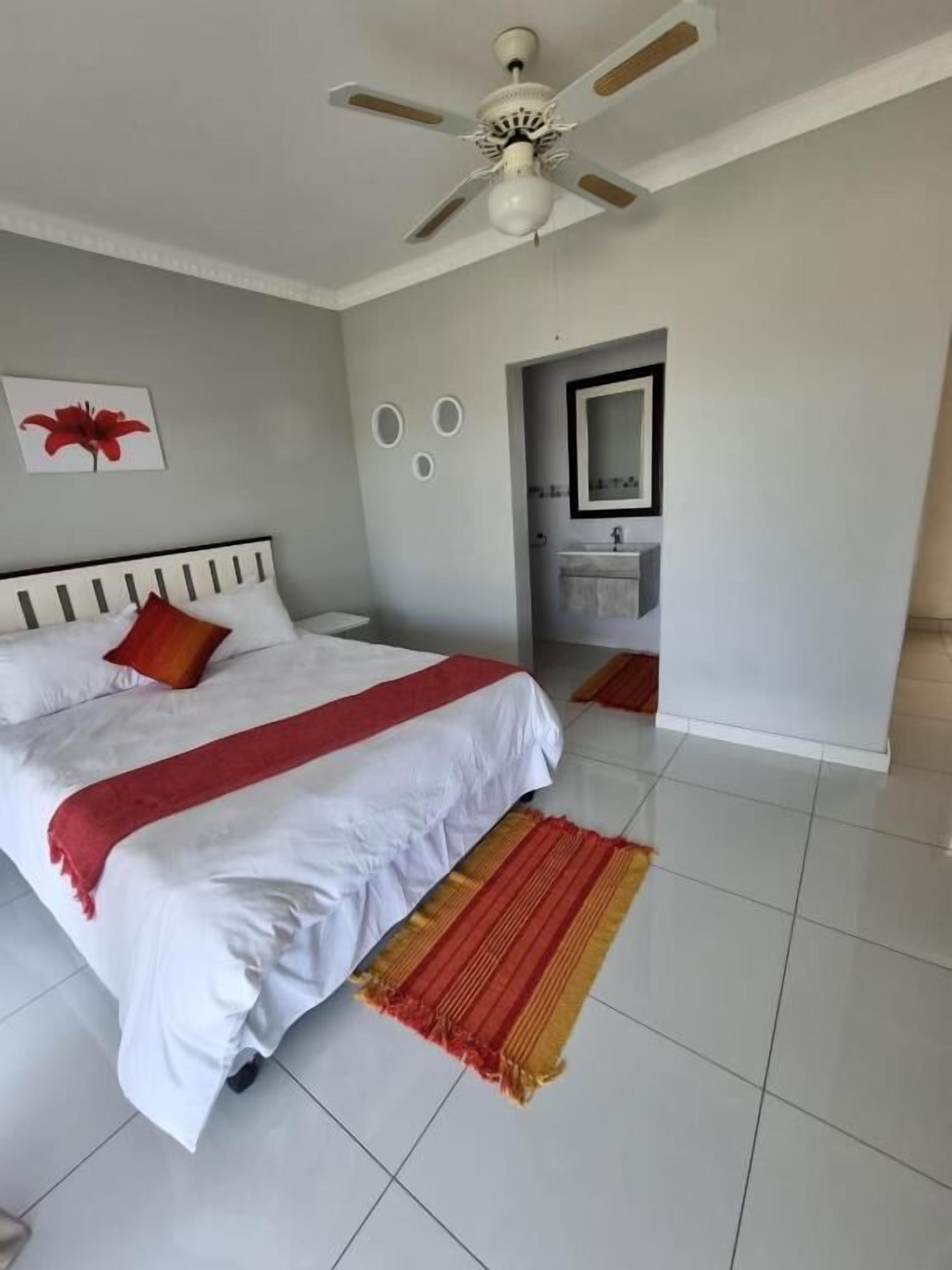 Quarto de hotel com cama, ventilador de teto, banheiro com espelho e um quadro de uma flor vermelha na parede.