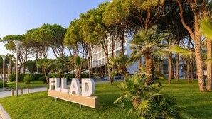 Placa com 'ELLAD', com um edifício moderno ao fundo, rodeado por vegetação exuberante e palmeiras.
