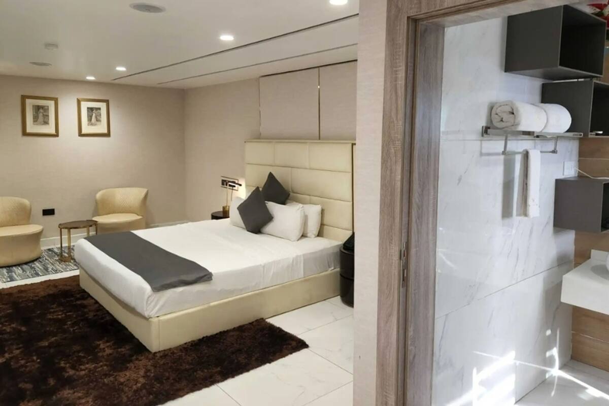 Quarto moderno com uma cama grande, duas poltronas, uma mesinha de apoio e cabeceira com iluminação embutida.