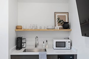 Uma cozinha compacta com bancada branca, pia de aço inoxidável, micro-ondas, cafeteira e prateleira com copos e uma moldura com uma fotografia.