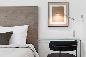 Uma cama bem arrumada com roupa de cama branca e cinza, uma cabeceira de madeira, uma obra de arte abstrata emoldurada, um abajur moderno e uma mesa lateral de vidro com uma cadeira preta.