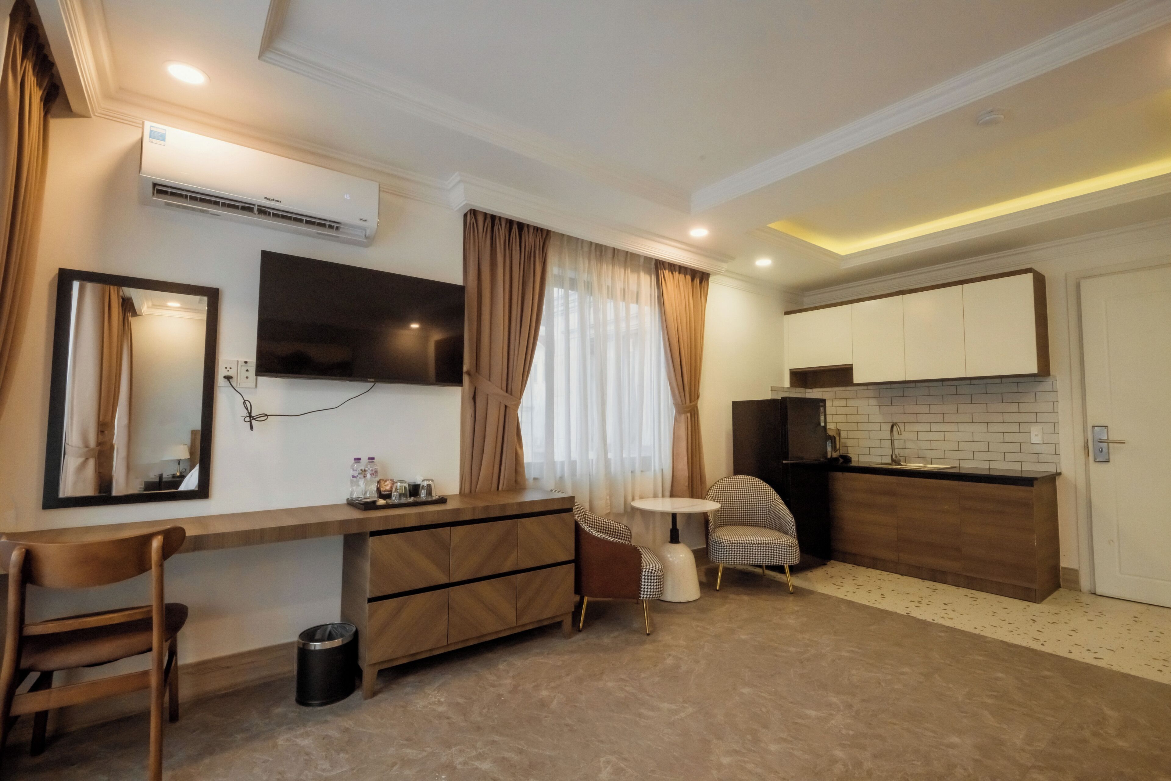 Quarto de hotel moderno com uma escrivaninha de madeira, uma cadeira, uma mesinha redonda, uma TV fixada na parede, uma cozinha com armários e uma lixeira.