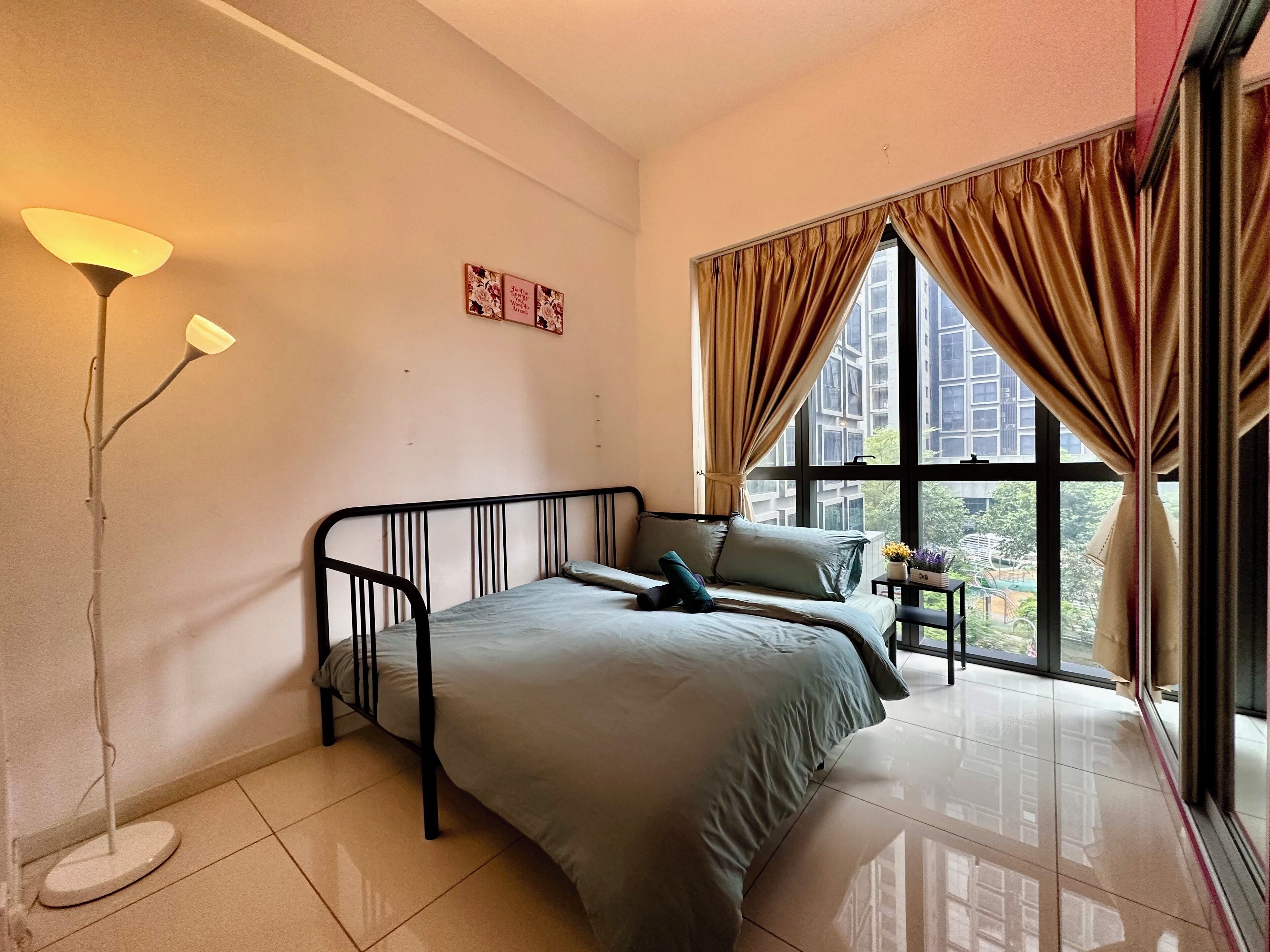 Quarto com cama, luminária de chão, janela com cortinas e vista para edifícios.