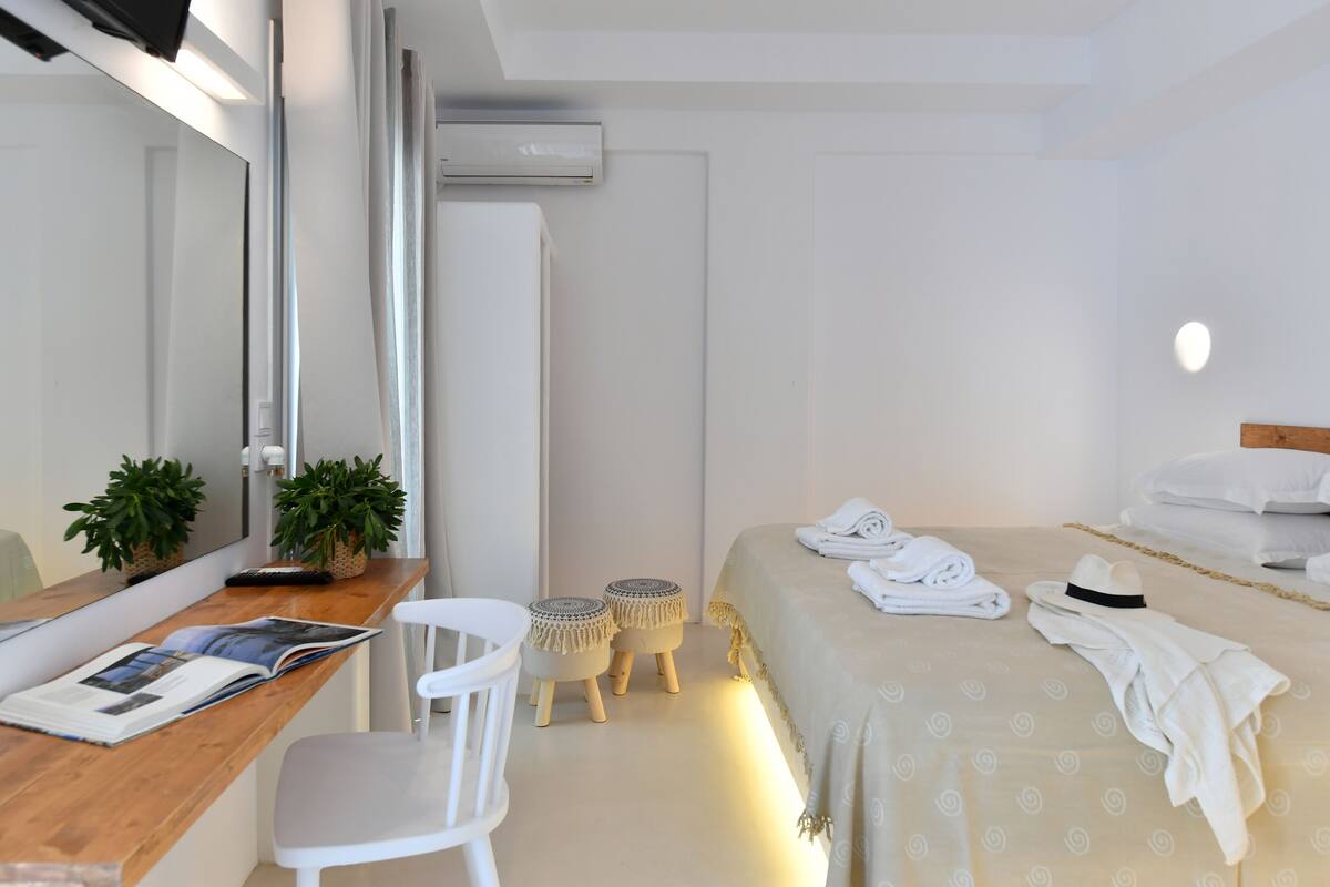 Quarto de hotel moderno com uma escrivaninha de madeira, dois banquinhos pequenos, uma cama com toalhas dobradas, uma cadeira branca e duas plantas em vasos.