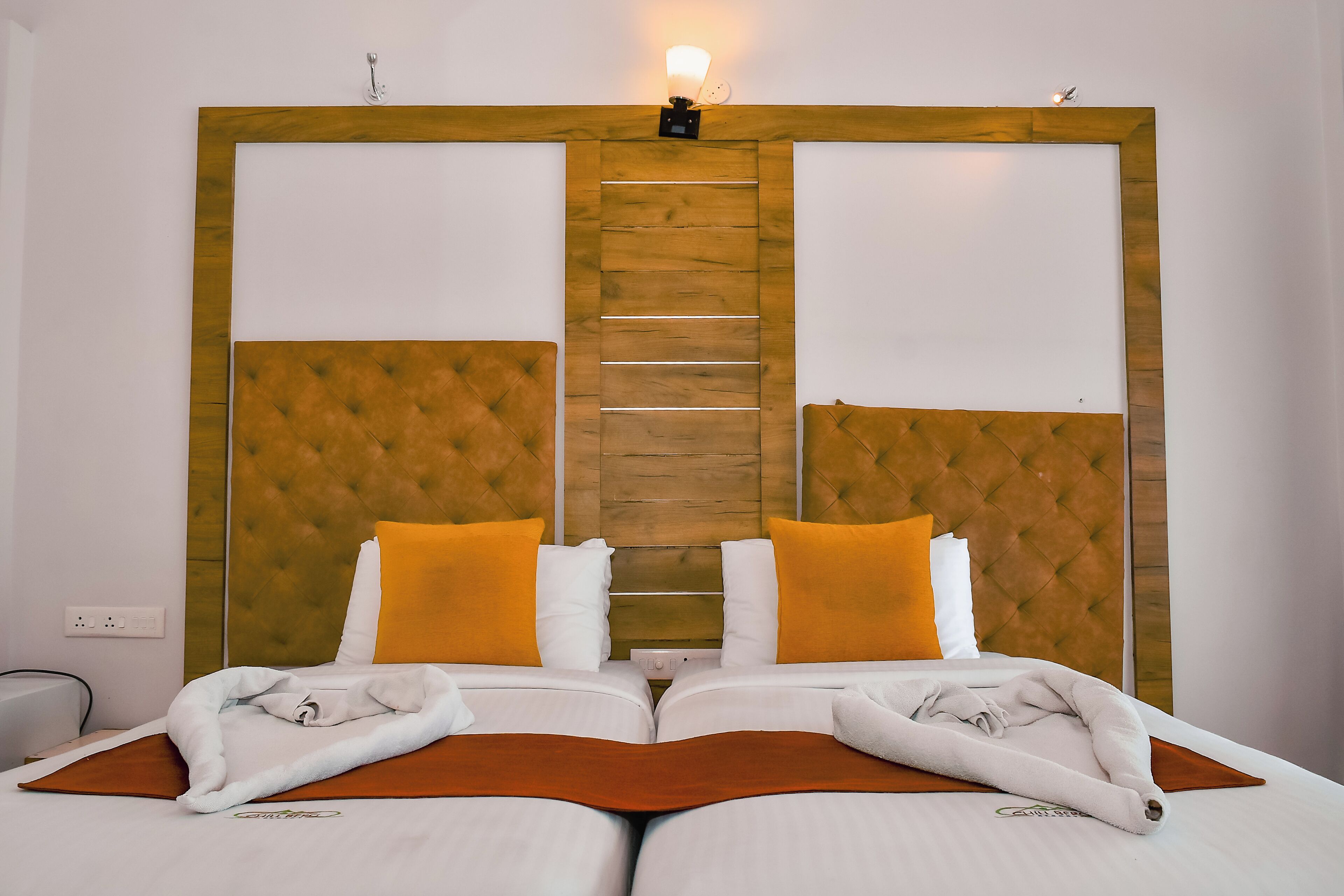 Uma cama de casal com cabeceira de madeira, duas almofadas brancas e uma almofada marrom decorativa. Há uma mesa de cabeceira com uma luminária.