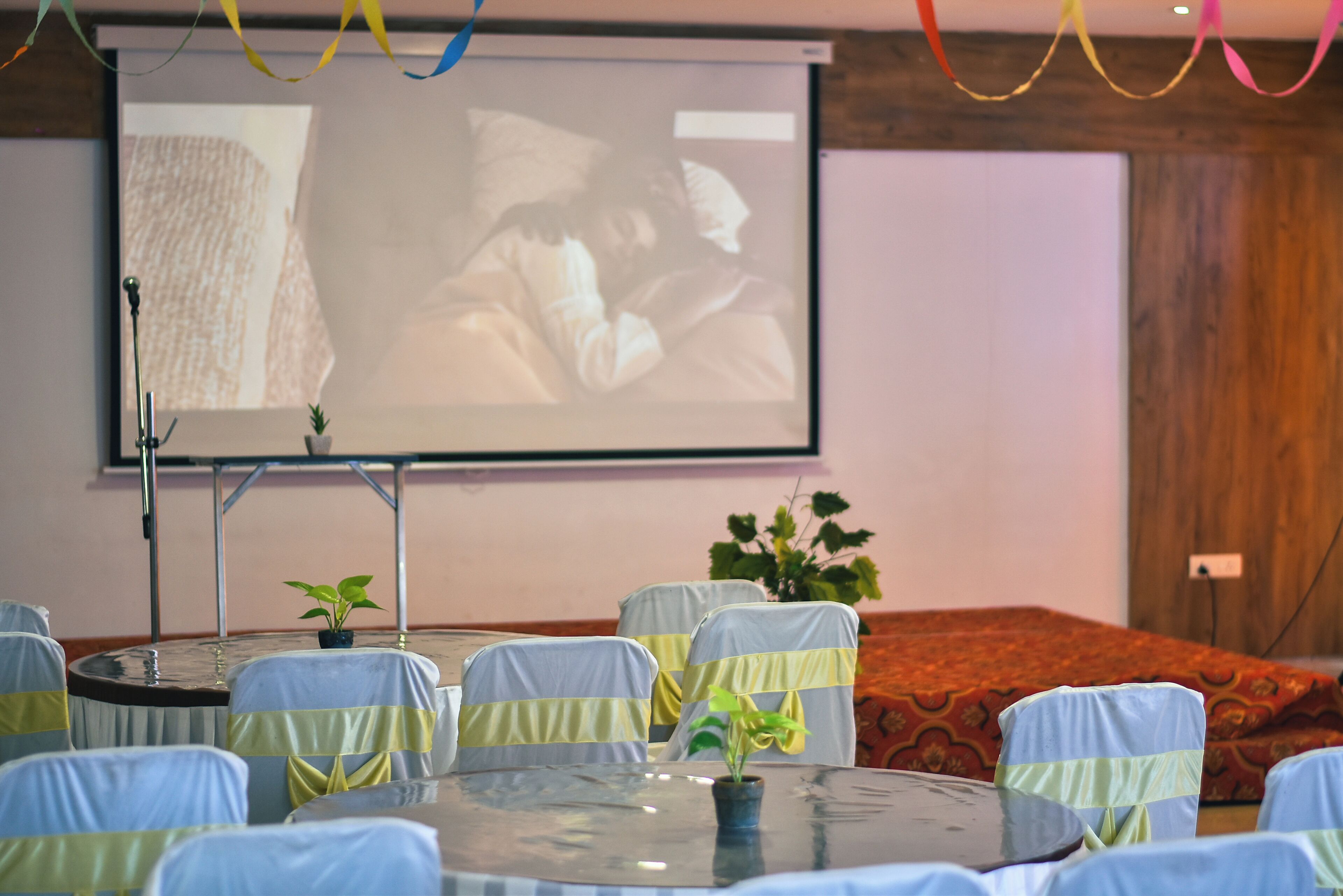 Uma sala de conferência com tela de projetor, mesas redondas, cadeiras com fitas amarelas e um vaso de planta.
