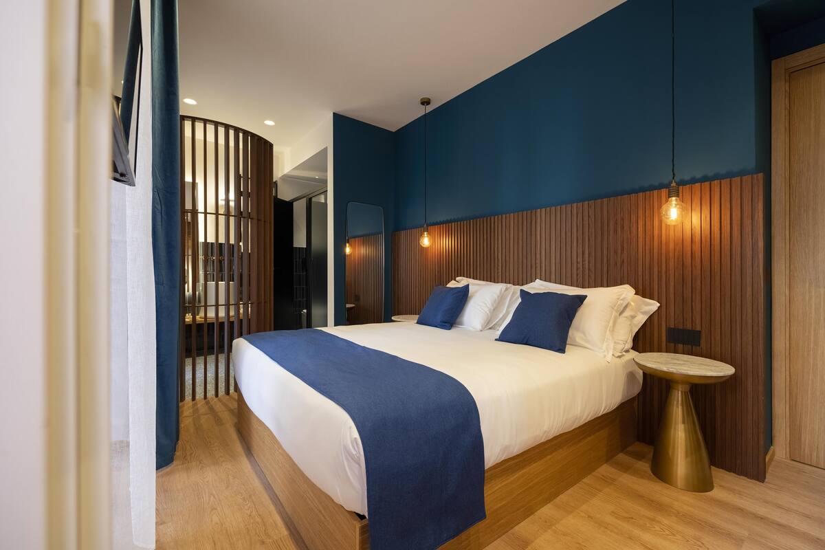 Quarto de hotel moderno com uma cama grande, uma mesa lateral redonda e uma parede de destaque em madeira.