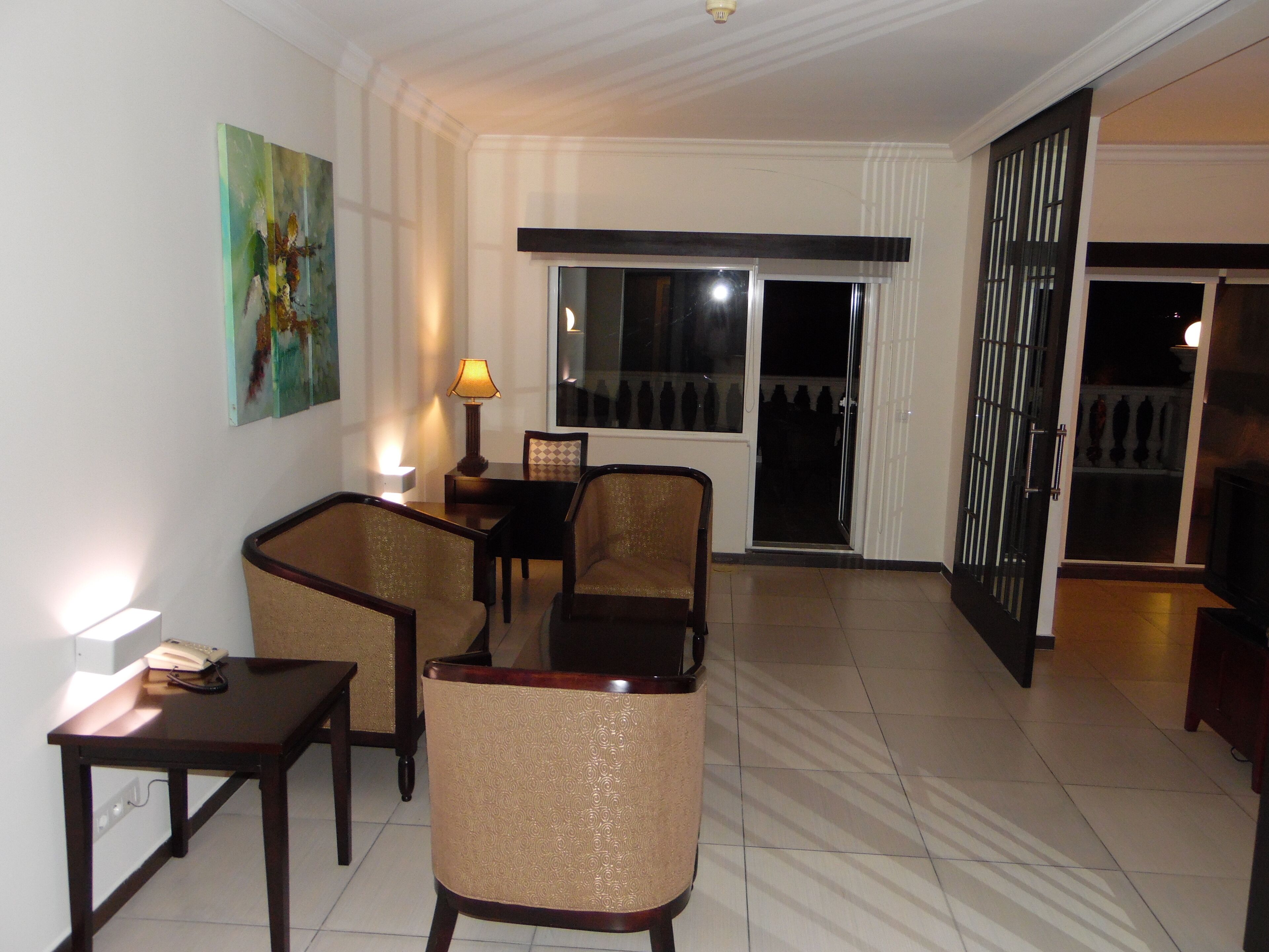 Quarto de hotel com área de refeições, sofá, mesa e luminária. Há uma porta de correr que dá para uma varanda.