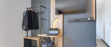 Quarto de hotel moderno com televisão de tela plana na parede, um laptop numa mesa de madeira, uma cadeira, um cabideiro e um móvel para guardar sapatos.