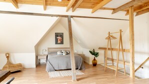 Quarto com teto inclinado, cama com manta cinza, escada de madeira e um cabideiro suspenso.