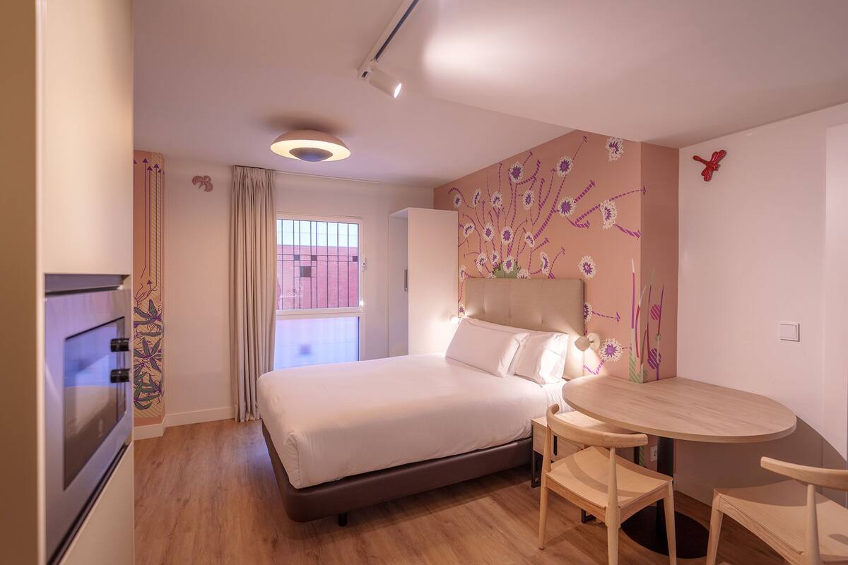 Quarto de hotel com cama, mesa, cadeiras, janela com cortinas e decoração floral e de borboletas na parede.